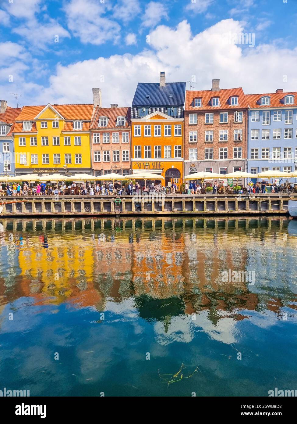 Colorful waterfront buildings reflect in calm water under a bright sky ...