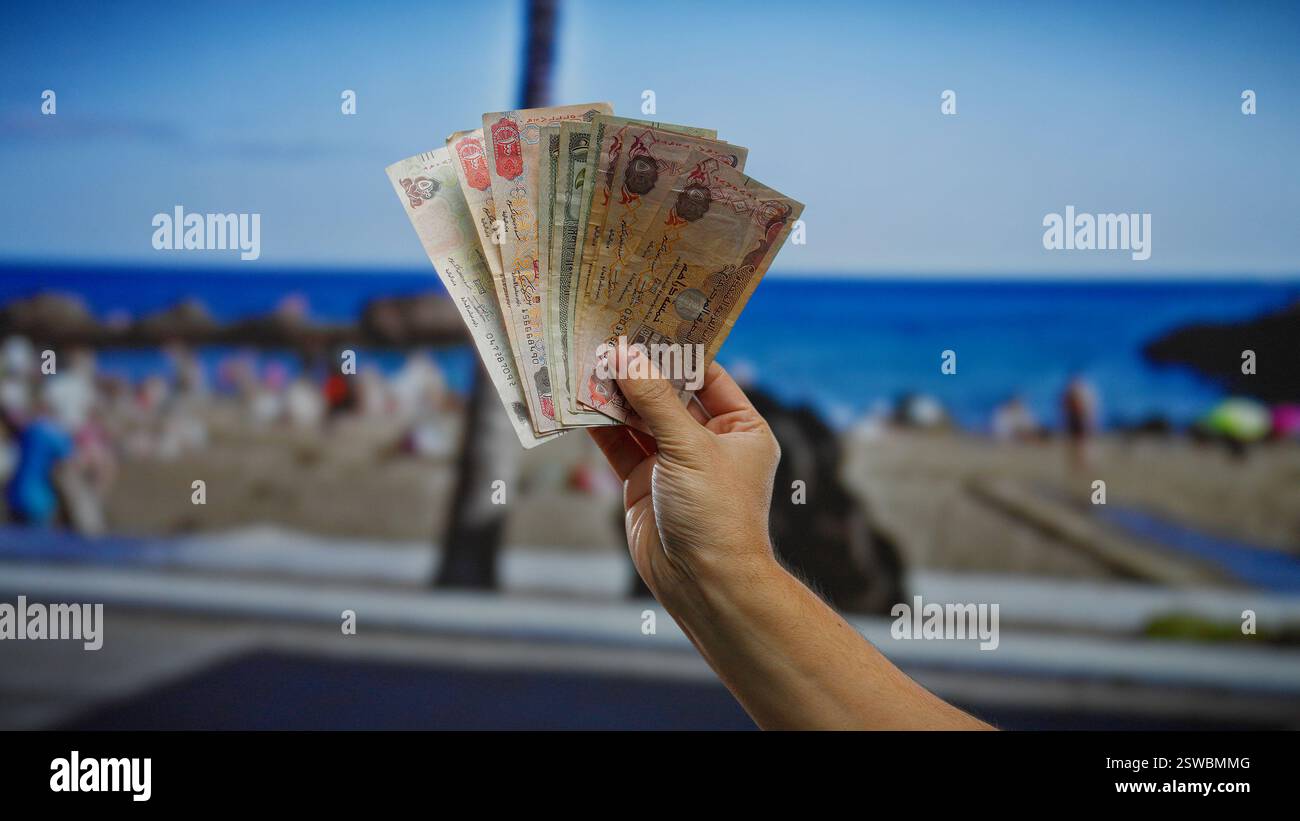 Hand holding emirati dirhams on a sunny beach background with people ...