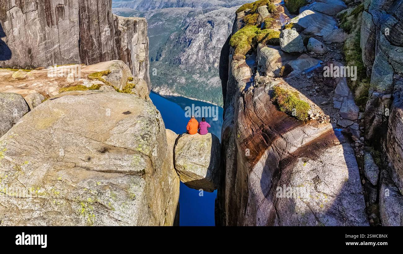 Two hikers sit on the edge of Kjeragbolten Pulpit Rock in Norway, enjoying the stunning ...