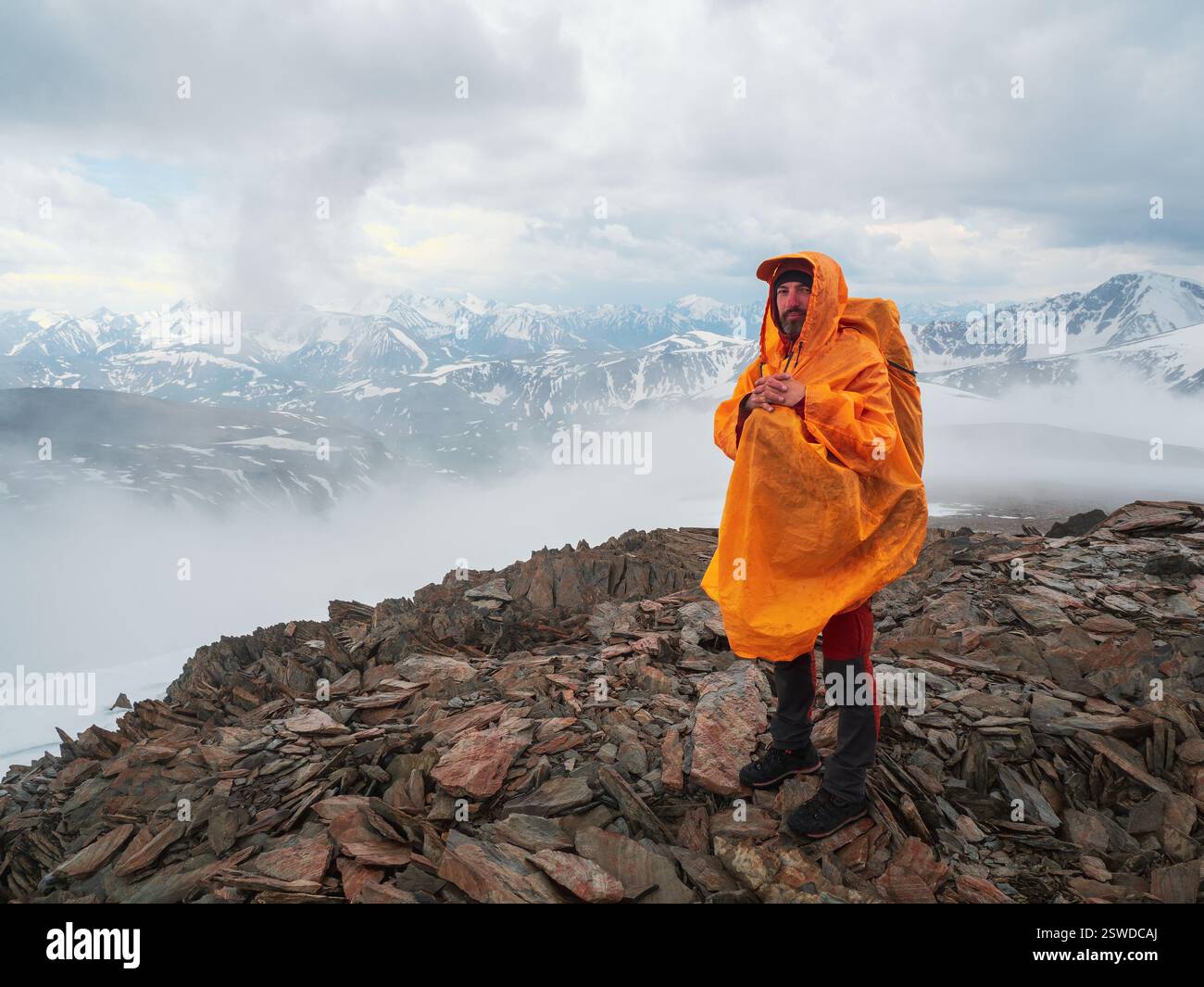 Tired tourist in a yellow raincoat on top of a mountain looks into ...