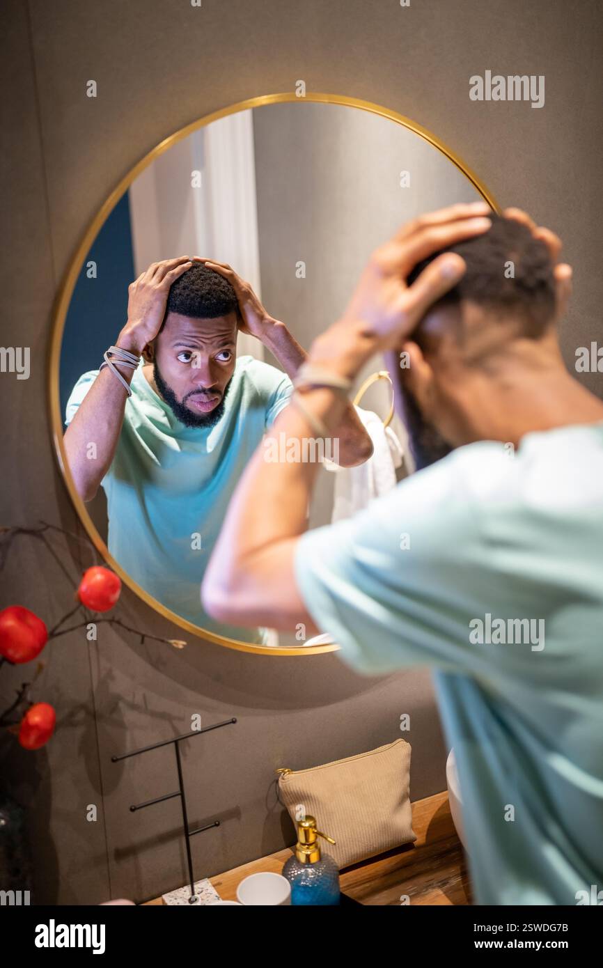 Upset young African American man looking in bathroom mirror touching ...