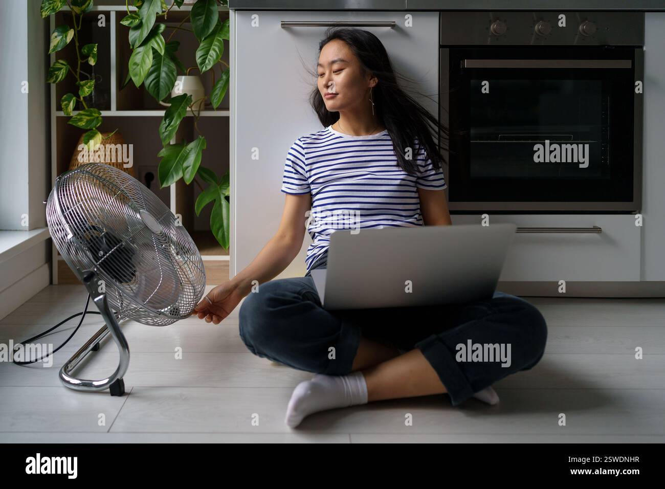 Happy smiling Asian girl freelancer sitting near electric fan working from home in heatwave ...