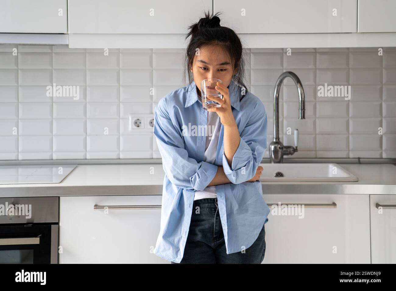 Unhappy disheveled Asian girl drinks water while standing in kitchen ...