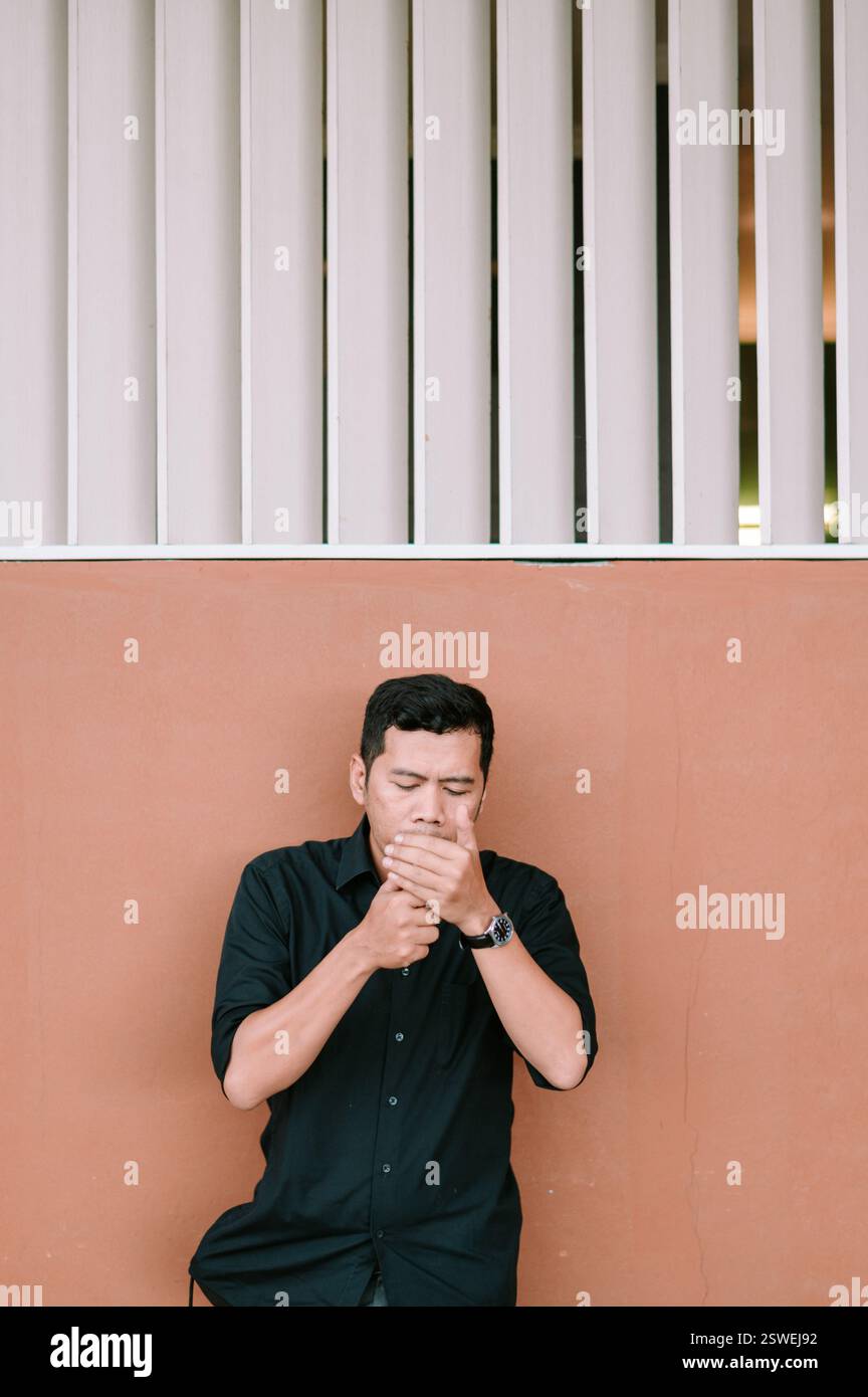Thoughtful young man standing against an orange wall, covering his ...