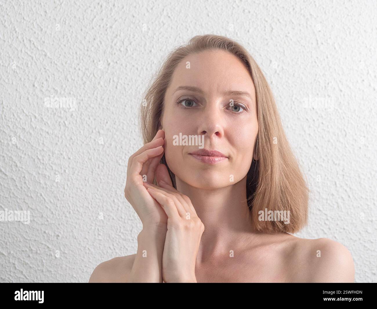 Portrait of beautiful middle aged woman touching her face on white wall. Beauty and skincare ...