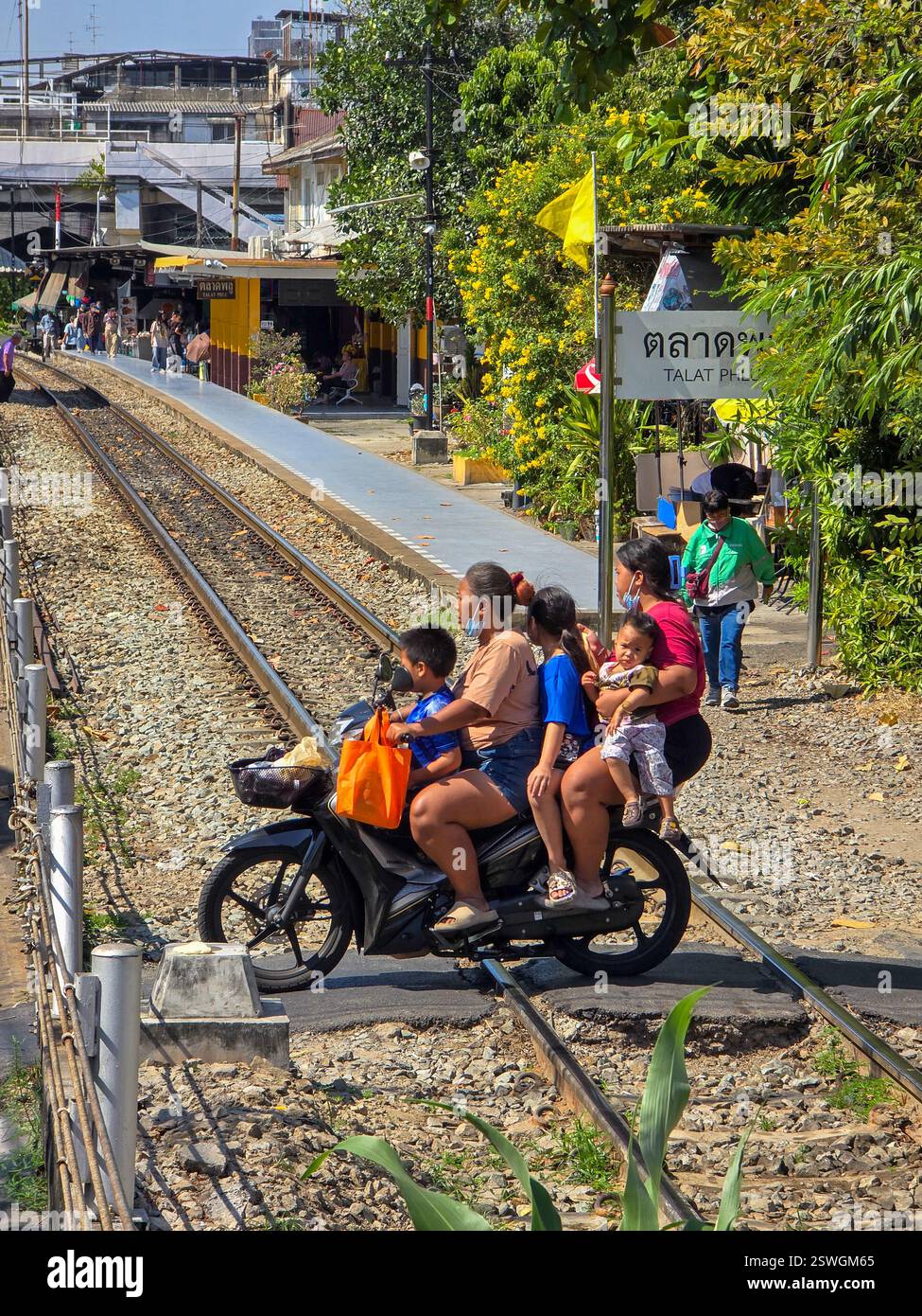 Family riding a scooter across train tracks in bustling Bangkok market ...