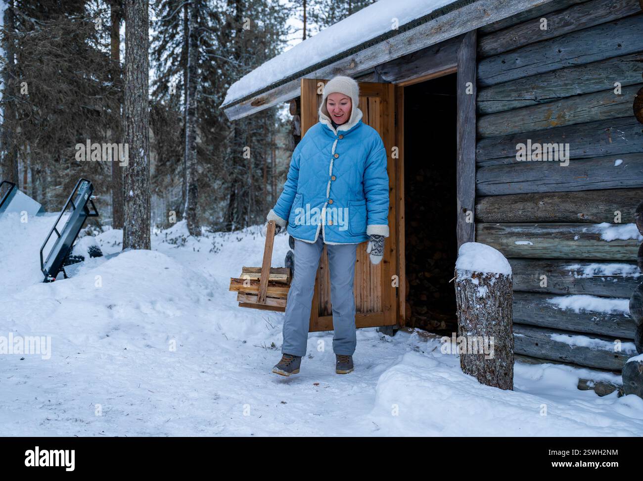 Woman in blue winter coat carrying firewood outside a rustic log cabin ...