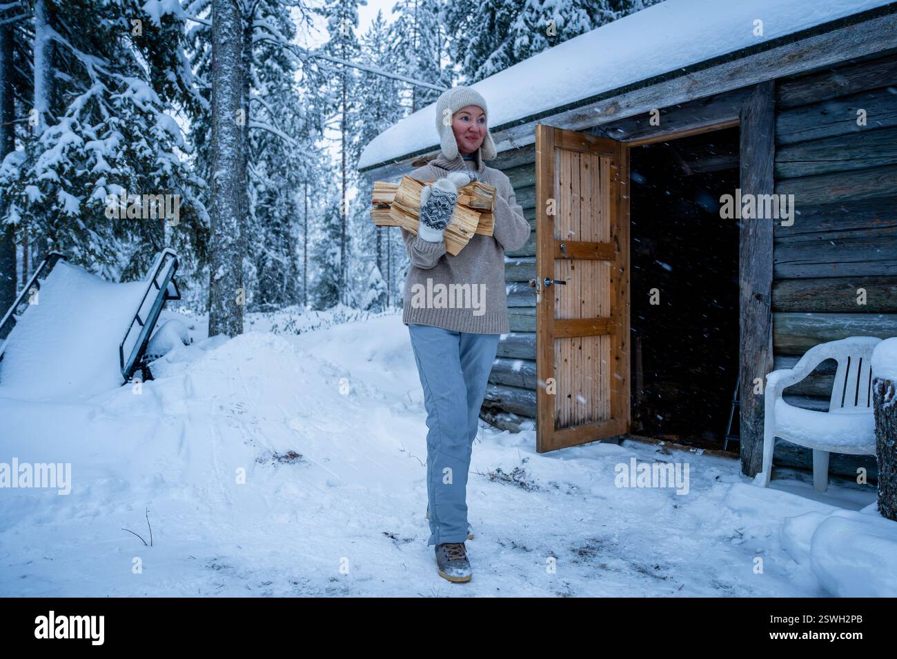 Woman in warm clothes carrying firewood outside a rustic log cabin ...