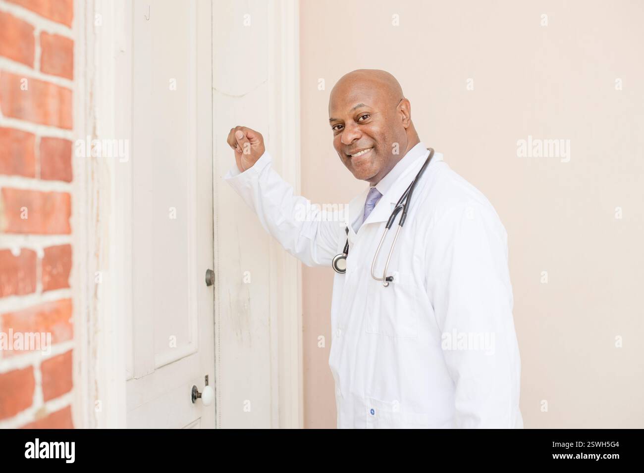 Smiling doctor in a white coat knocking on a door for a house call ...