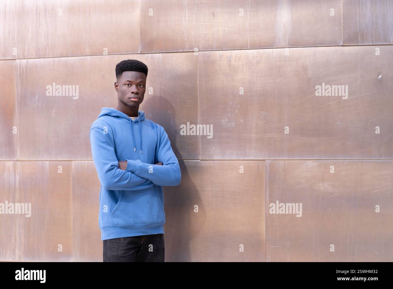 A young man in a blue hoodie stands in front of a wall. He is looking ...