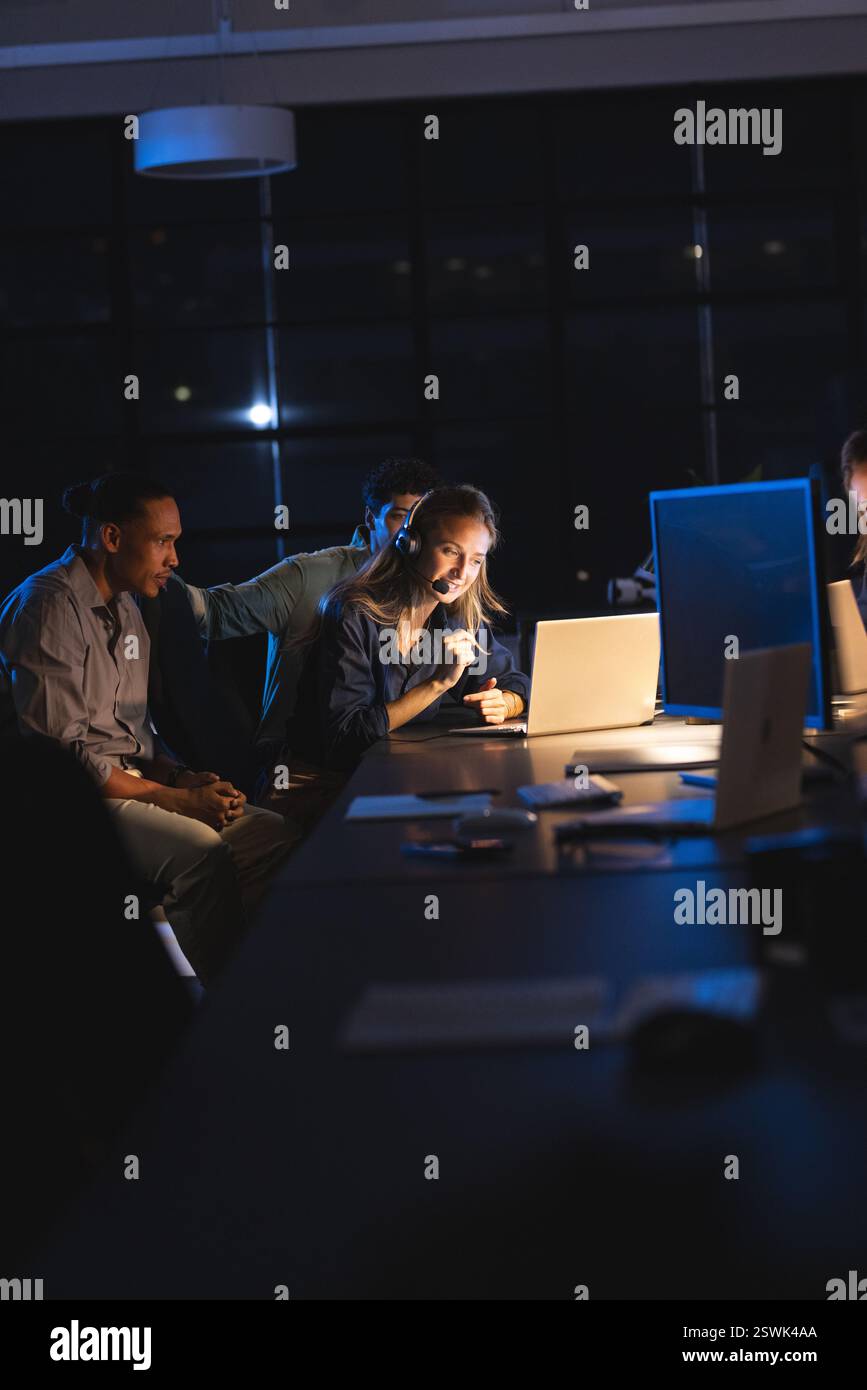 Diverse team collaborating at night in office, focused on laptop ...