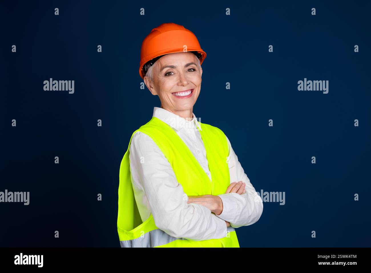 Confident senior businesswoman in construction attire standing before a dark blue background ...