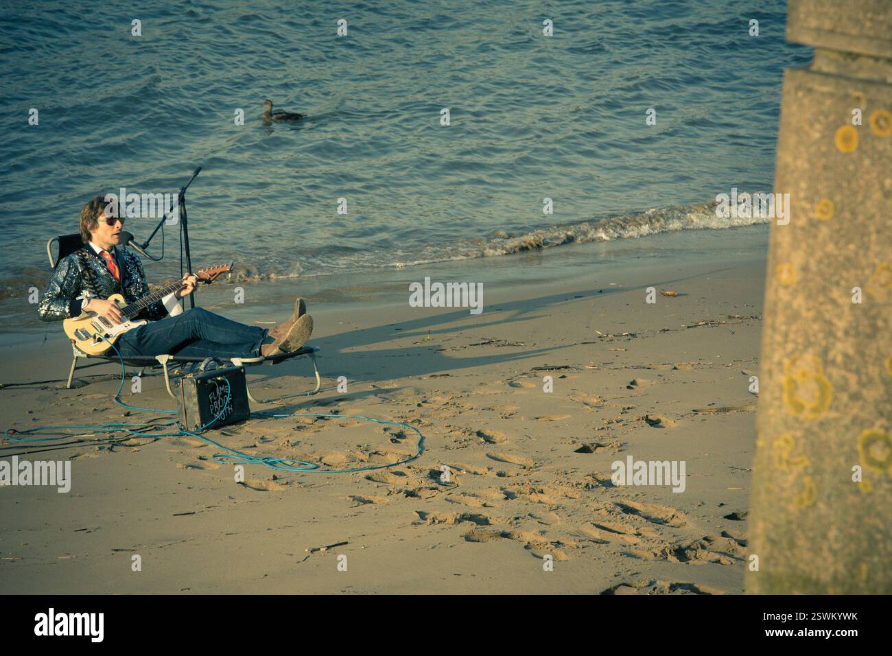 A musician plays guitar on the beach near the Thames, London. Waves lap ...