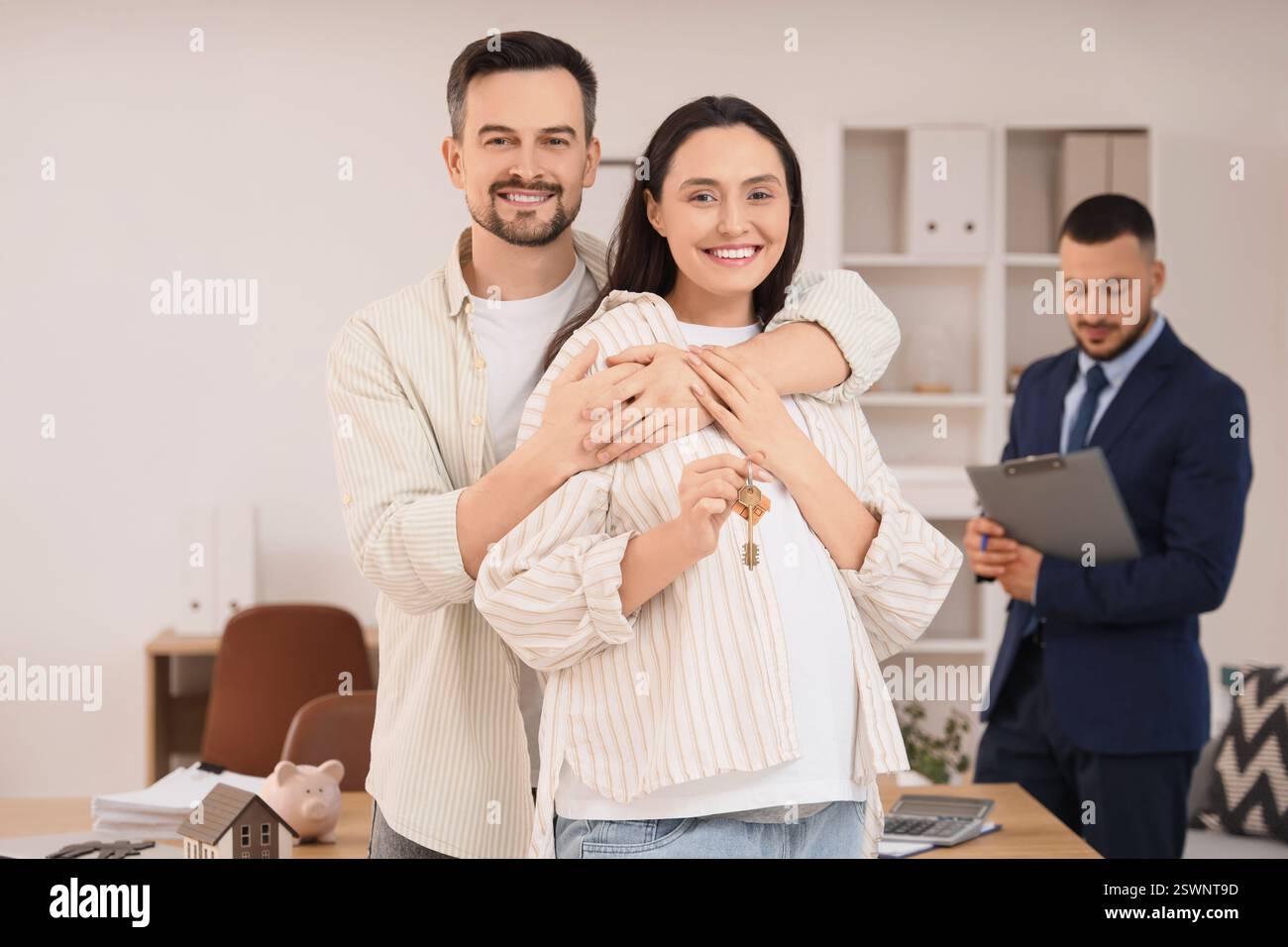 Young pregnant couple with key from house hugging at real estate agent ...