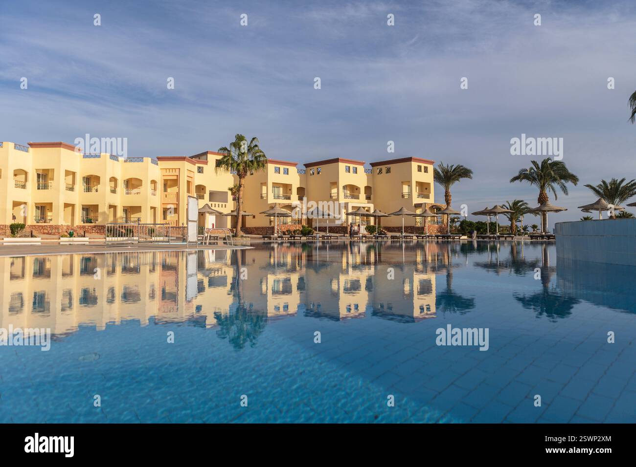 Palm trees, sun loungers, and a large resort hotel are reflected in a calm swimming pool in ...