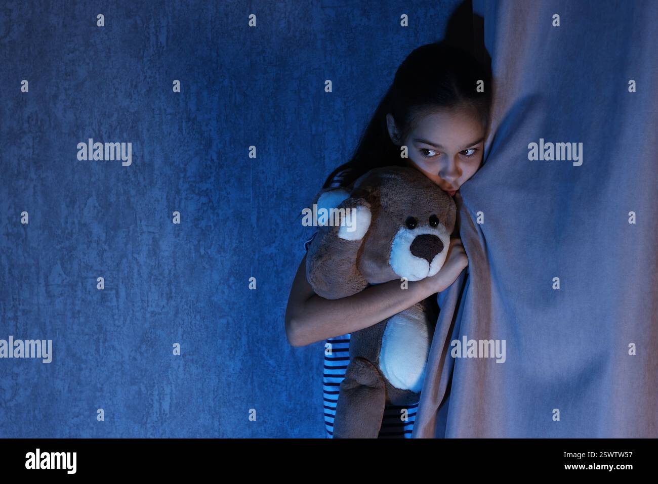 Scared girl hiding with teddy bear behind curtain at night. Space for ...