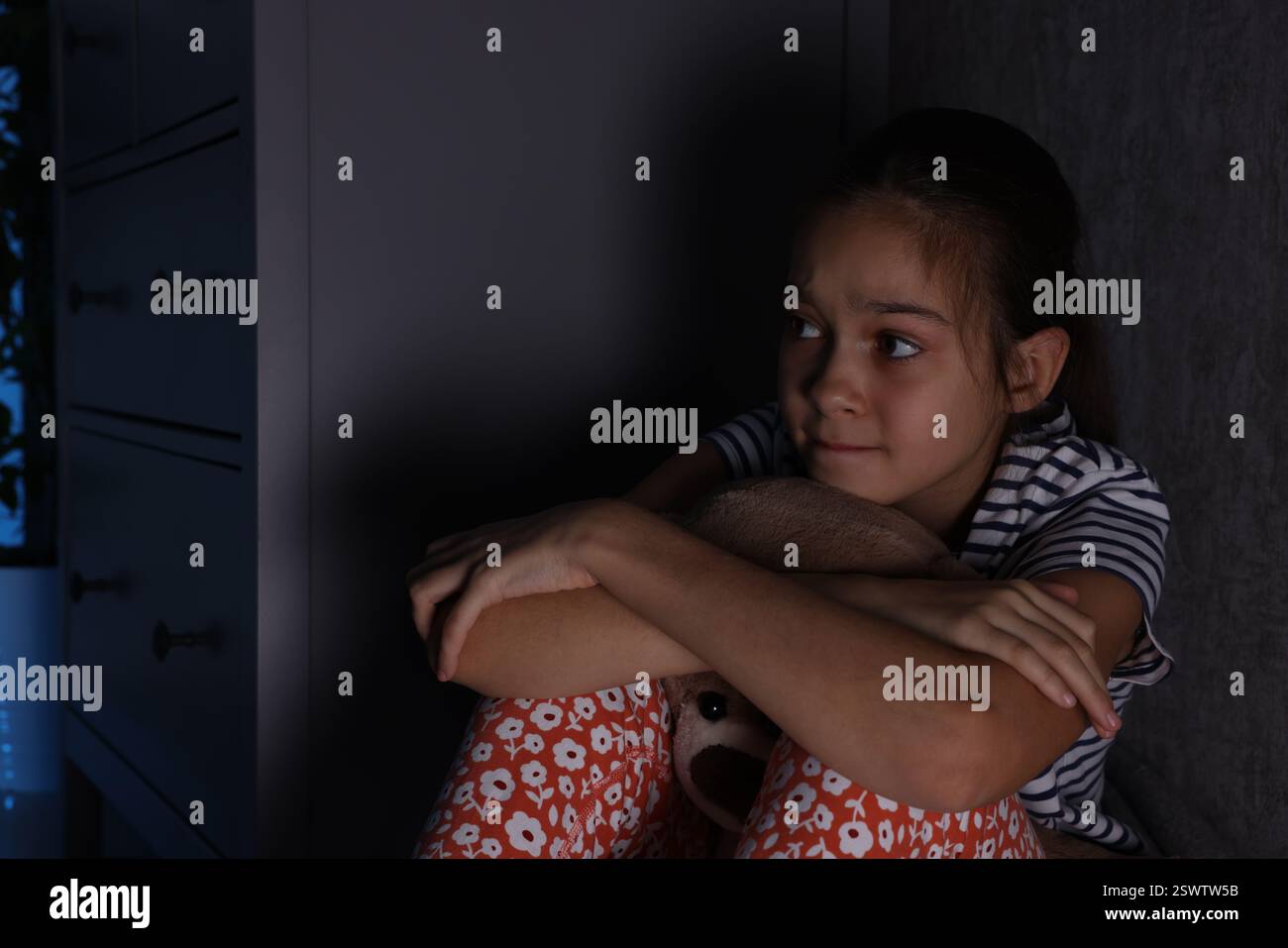Scared girl with teddy bear hiding behind chest of drawers in her room ...