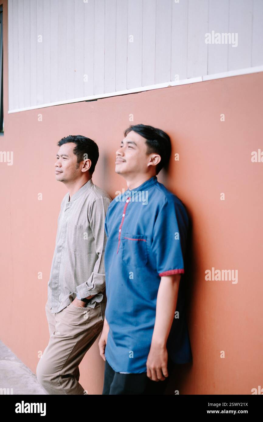 Two asian men in casual attire stand side-by-side against a coral wall ...