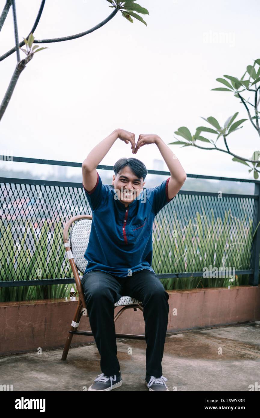 A young man sits on a terrace, making a heart shape with his hands. He ...