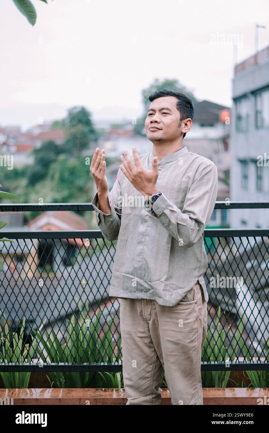 A young muslim man praying with raised hands on a balcony. He is ...