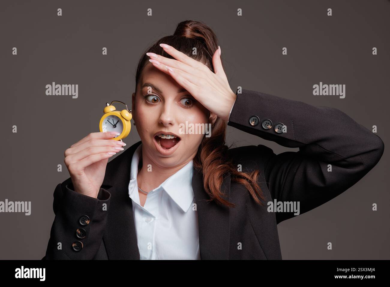 Stressed businesswoman in a suit holding a small alarm clock, shocked ...