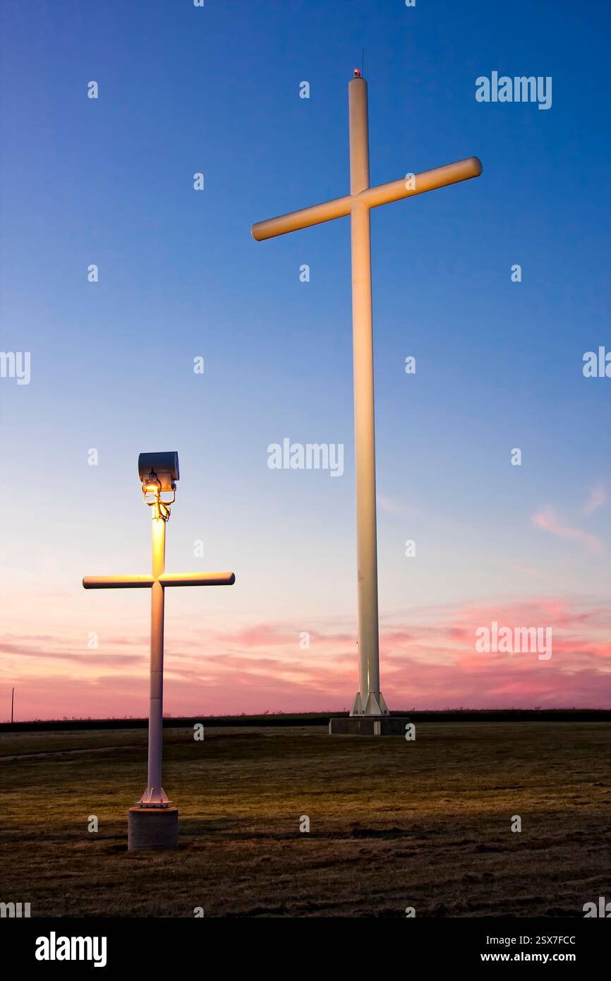 White cross is lit up against a blue sky. The cross is on a hillside ...