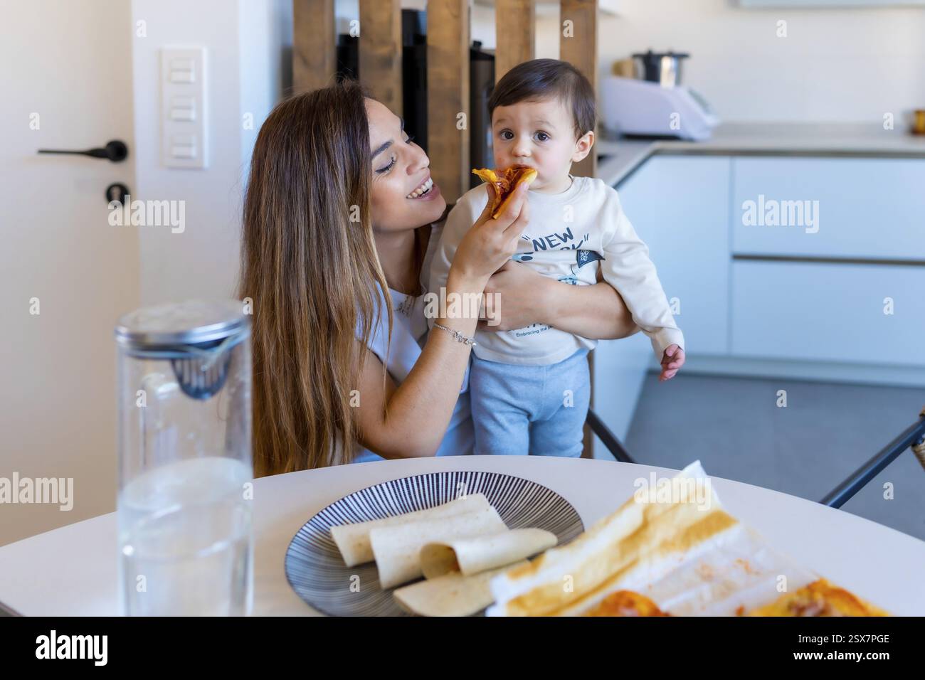 Young mother feeding her baby a slice of pizza while preparing mexican ...