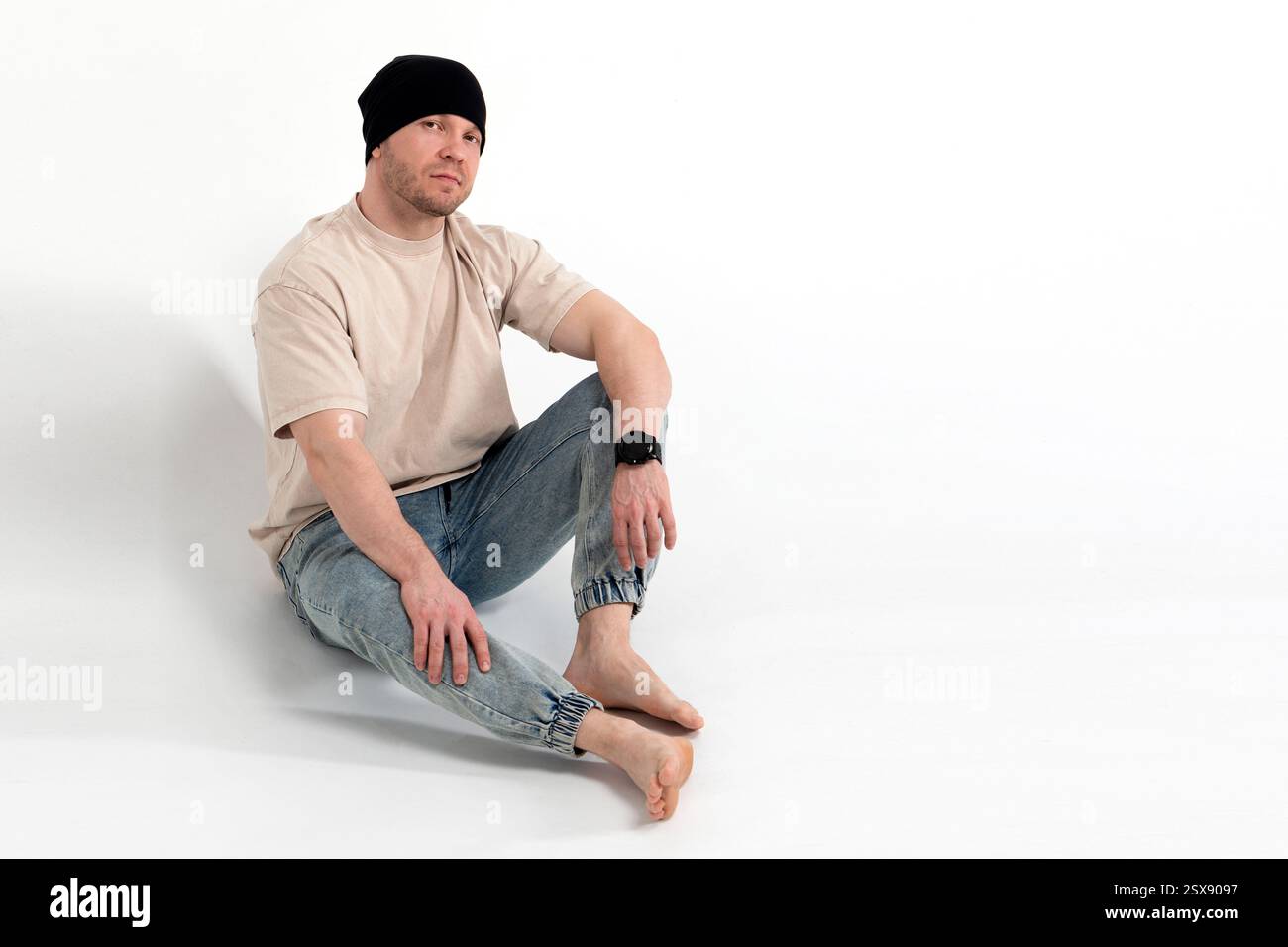 Man in beige t-shirt and black beanie sitting barefoot against white ...