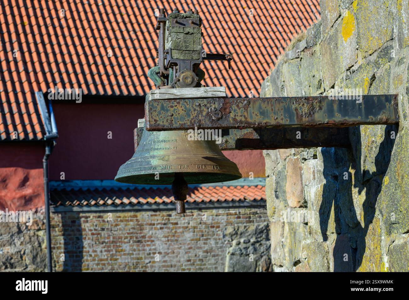 A bell hangs from a sturdy metal bracket attached to an aged stone wall ...