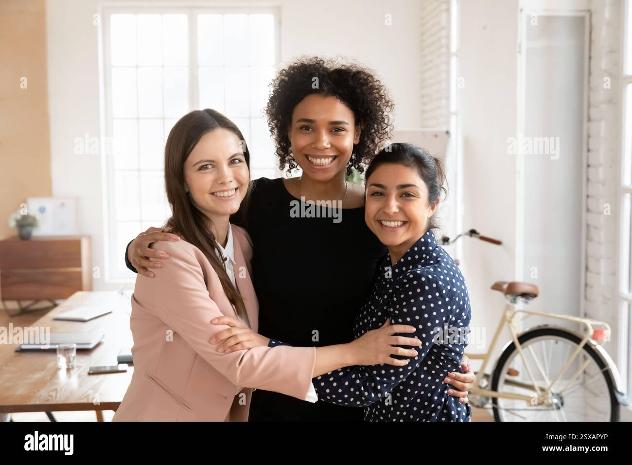 Three beautiful multinational women colleagues, hugging, looking at ...