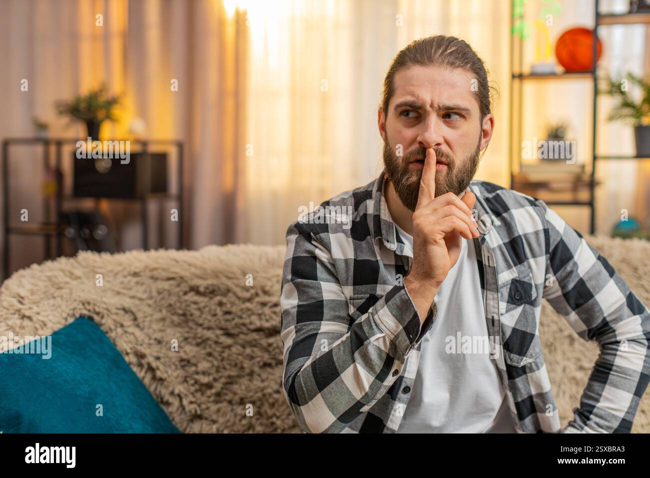 Young man sits at home with finger over mouth, whispering shush and ...
