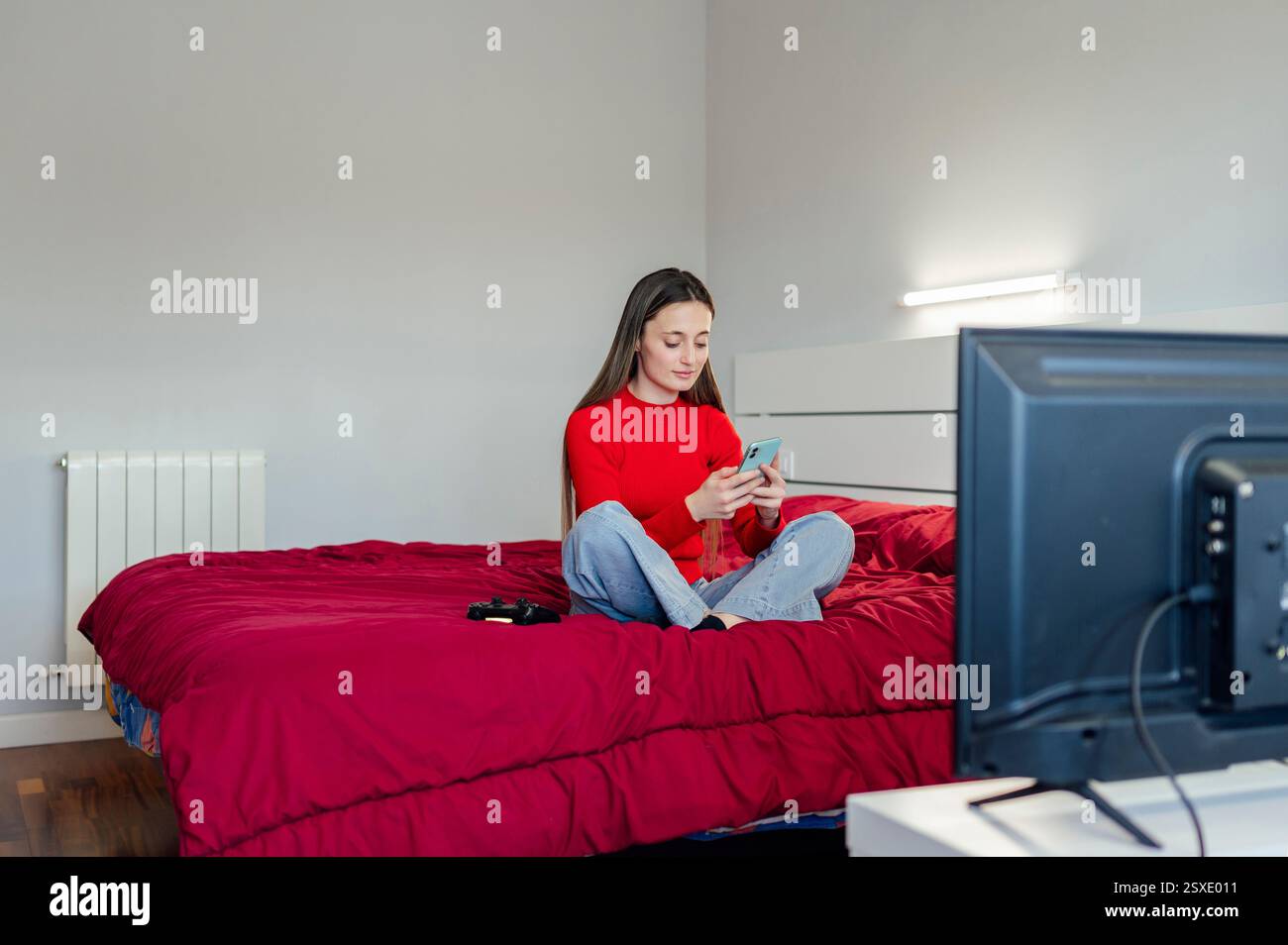 Woman using smartphone and holding controller while sitting on bed ...