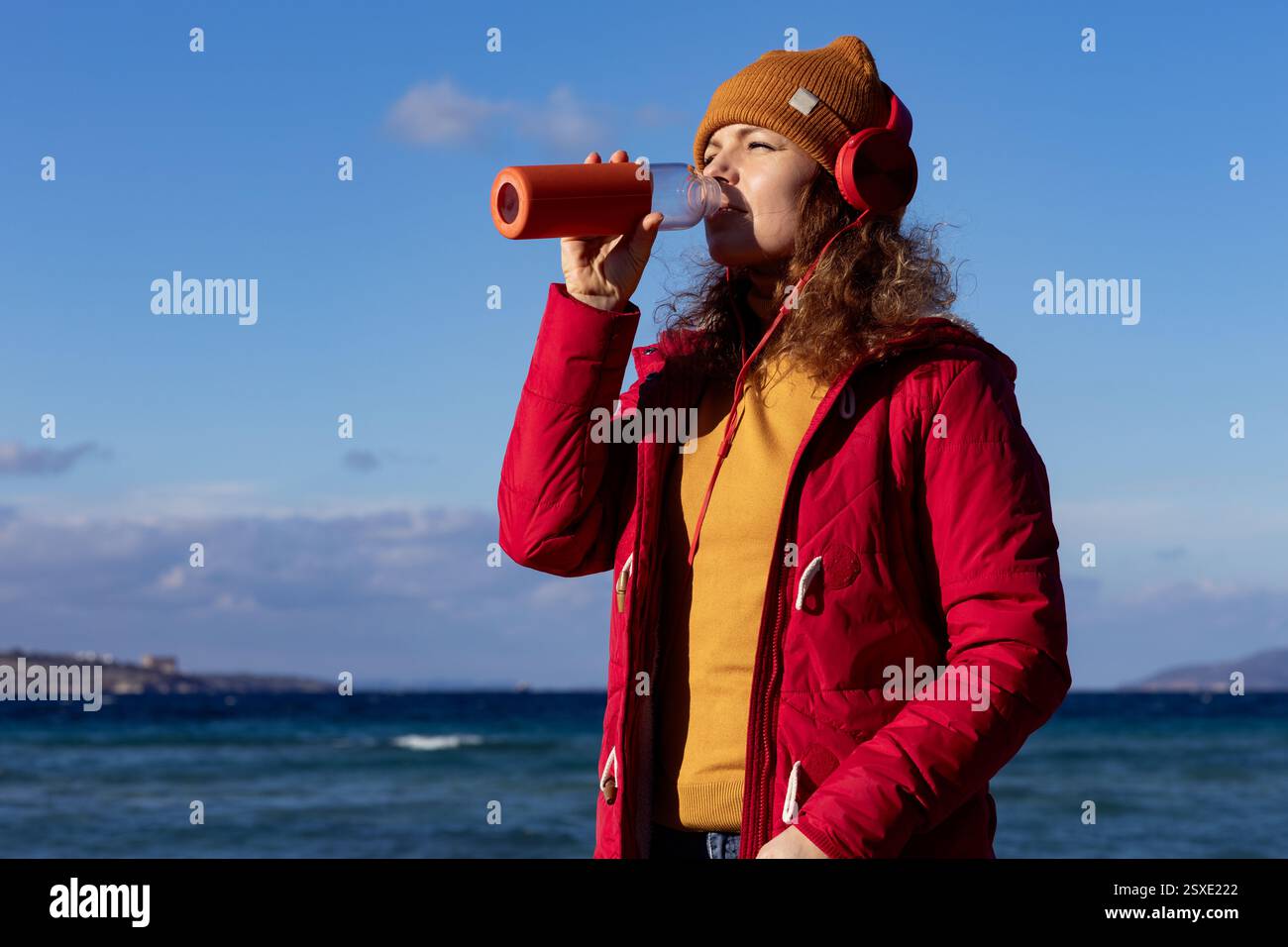 Cute woman with headphones drinking water by the sea in cold weather ...