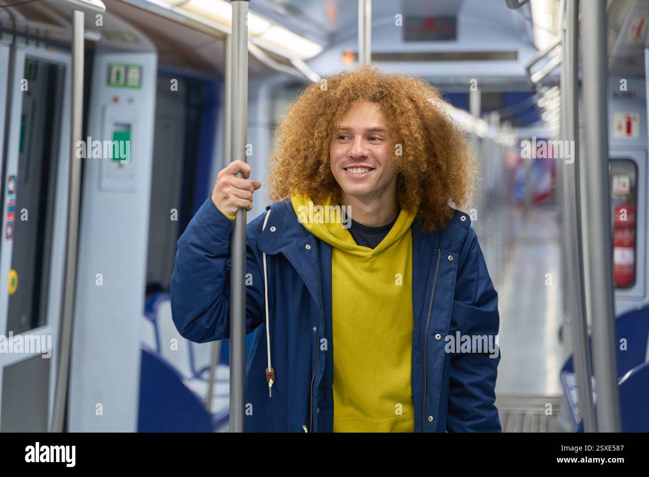 Smiling man with curly hair holding a pole in a subway train, enjoying ...