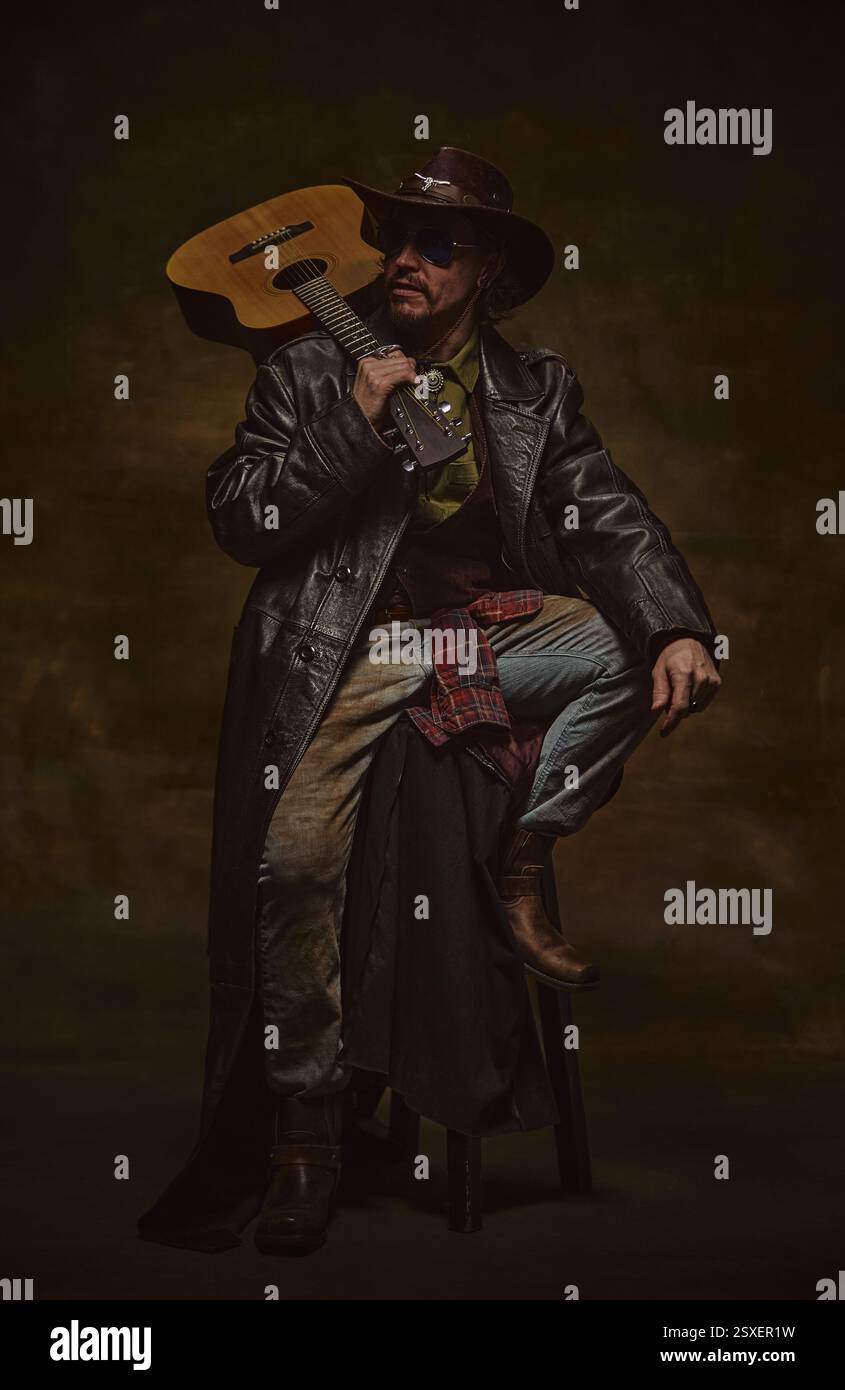 Brutal man, cowboy sitting on stool in leather coat, hat and boots ...