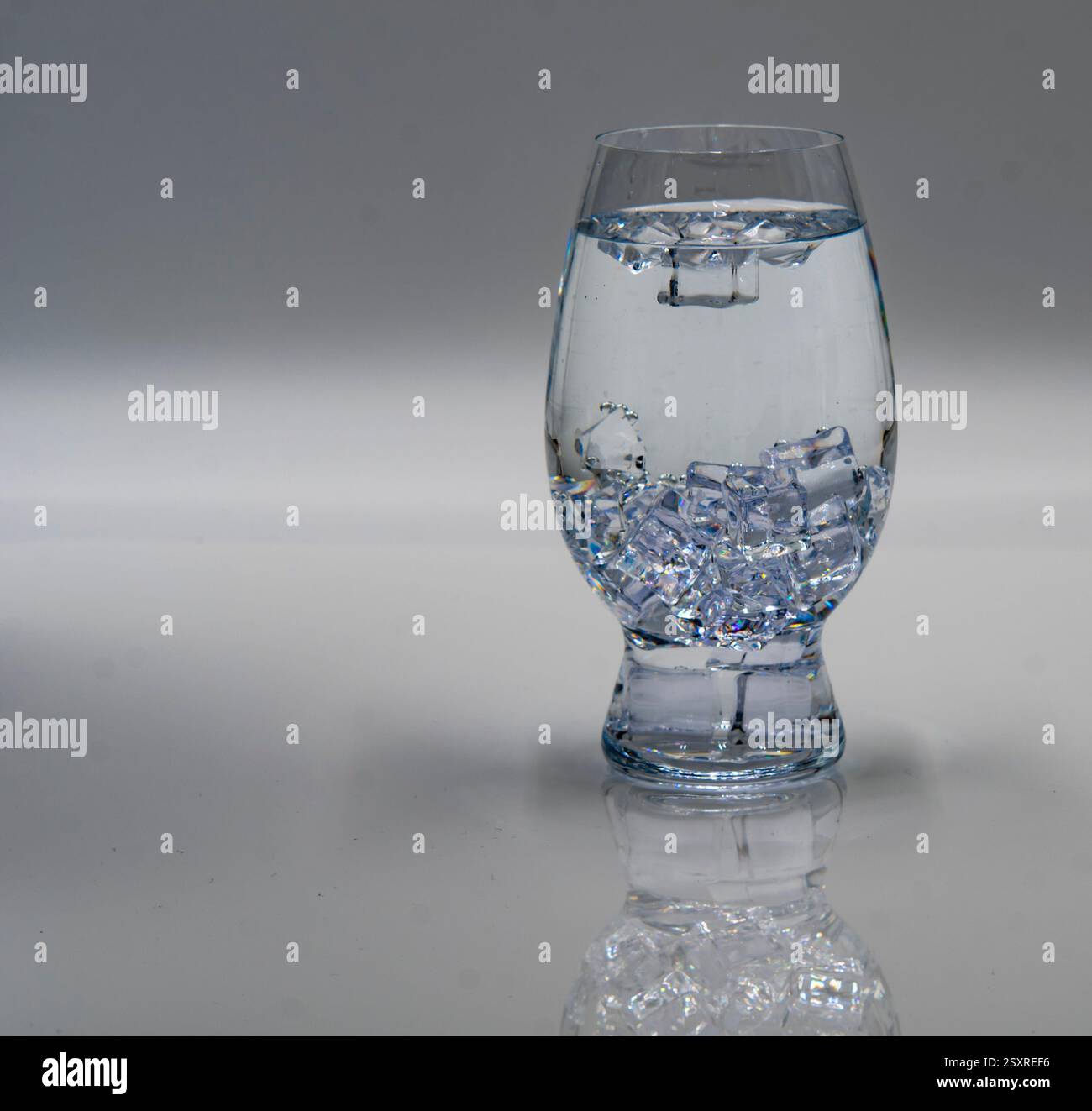 A clear glass filled with water and ice cubes sits on a smooth, reflective surface. The light ...