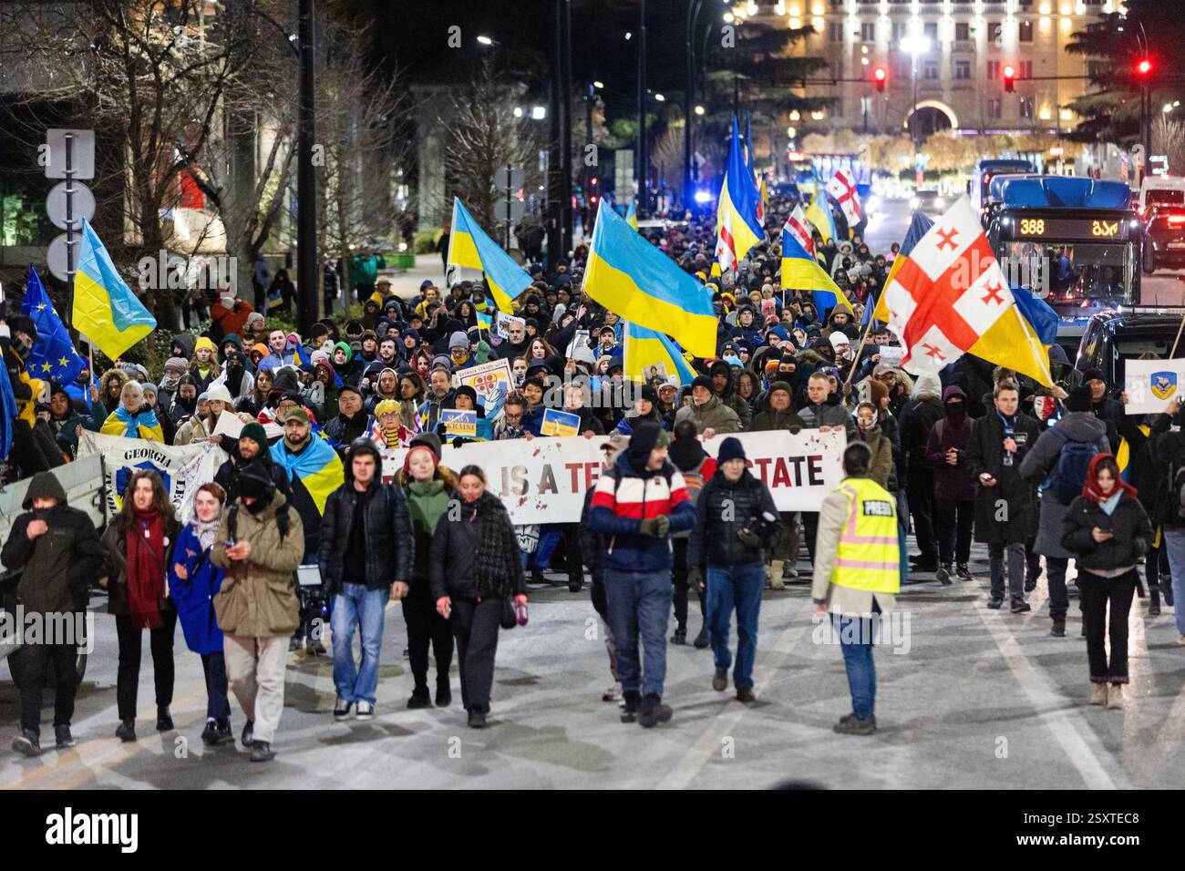 A large crowd of protesters marches down a brightly lit street in ...