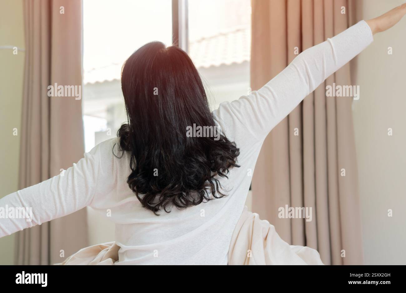Back view image of Asian woman is waking up in the morning, stretches in the bed, sun shines on ...