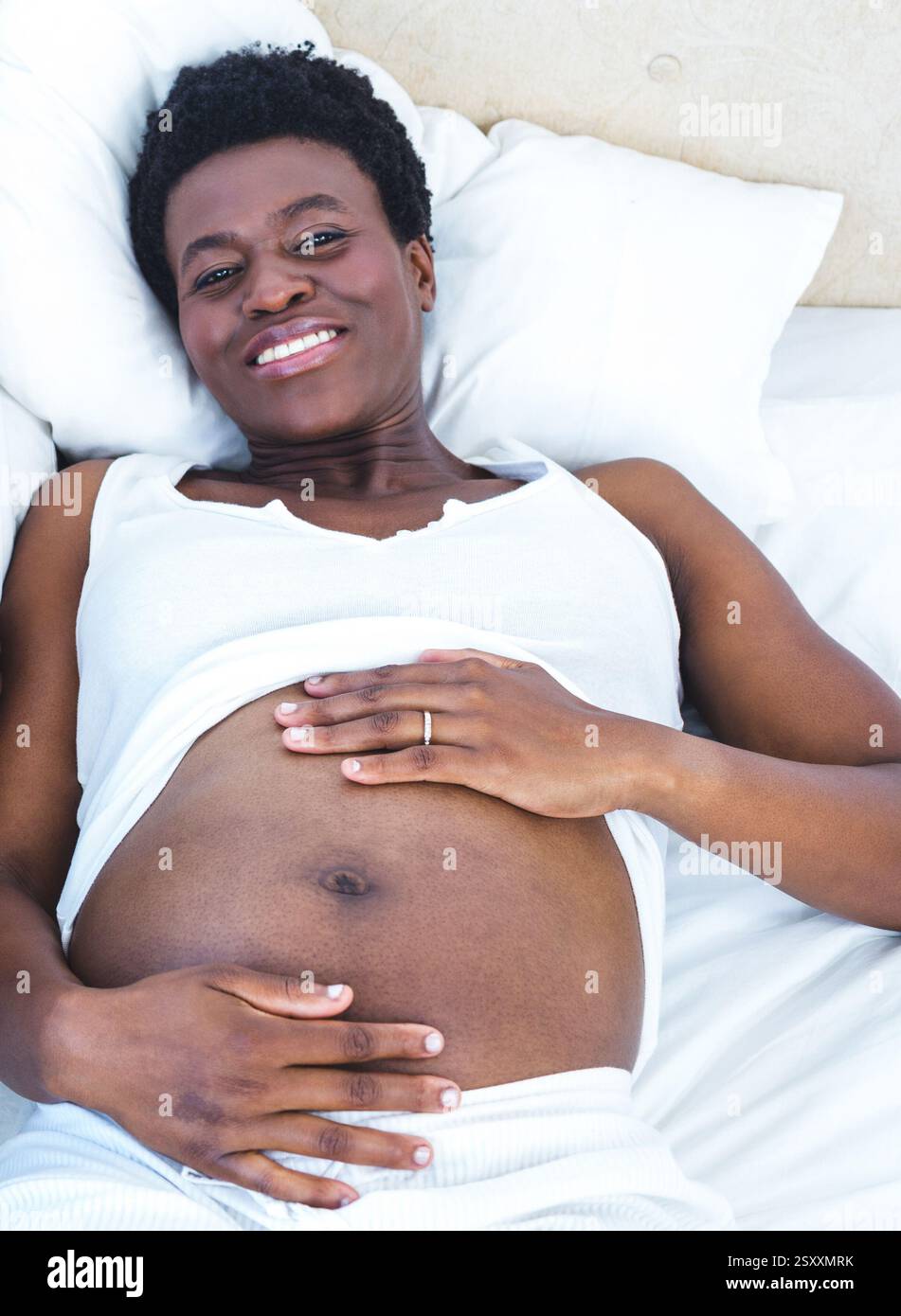 Pregnant woman relaxing on bed, smiling and embracing her belly with ...