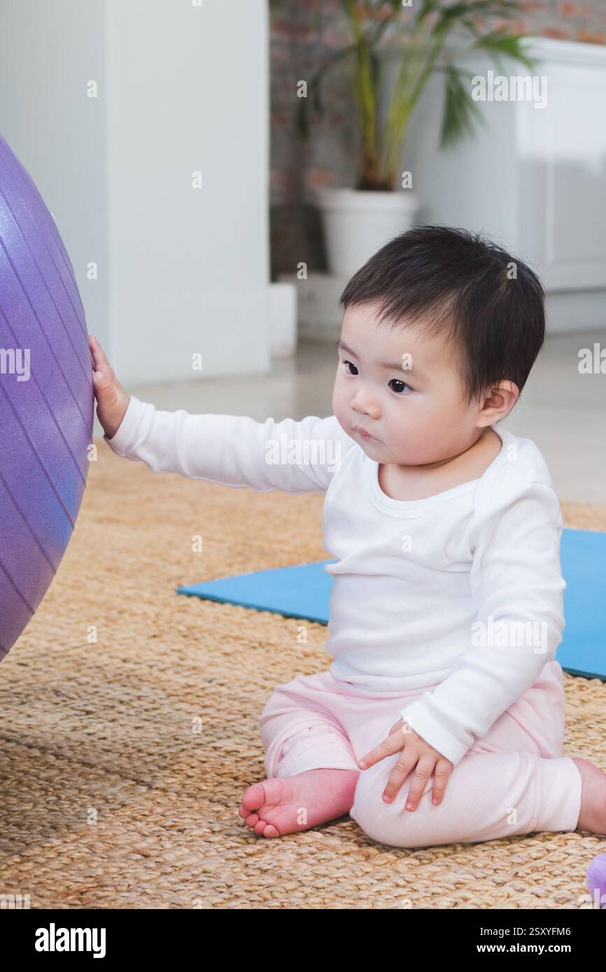 Asian baby playing with exercise ball on floor at home, looking curious ...