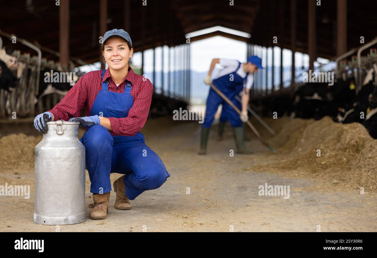 Young positive female farmer carrying milk churn near stall with cows ...