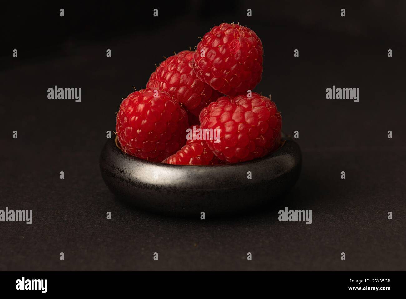 A small dark dish of fresh red raspberries , set as a still life on a ...