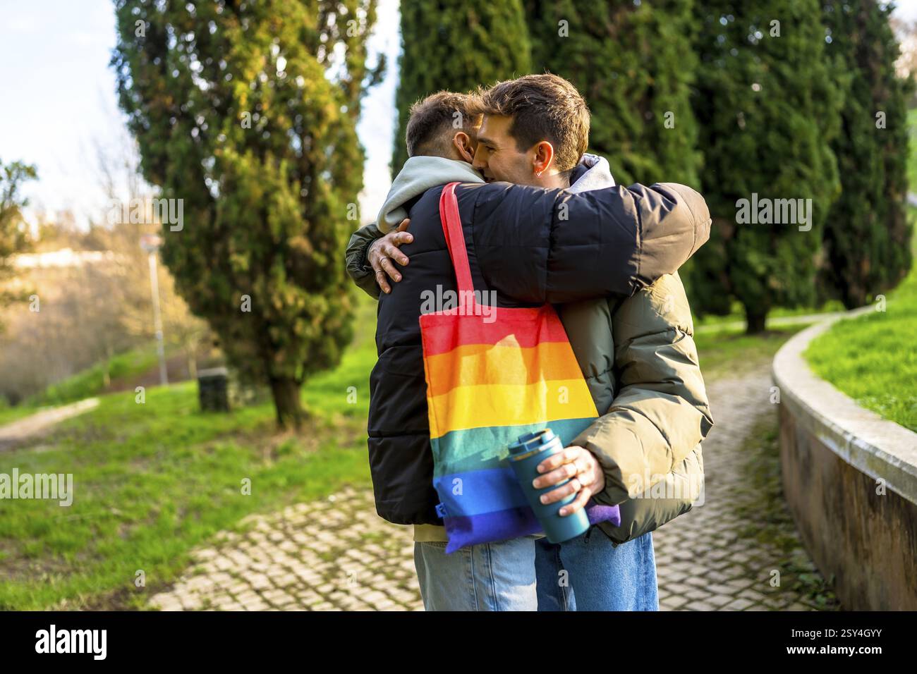 Two men embracing in a park, one carrying a rainbow tote bag ...