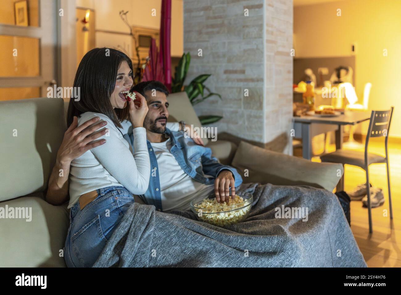 Young couple enjoying a movie night at home, eating popcorn from a bowl ...