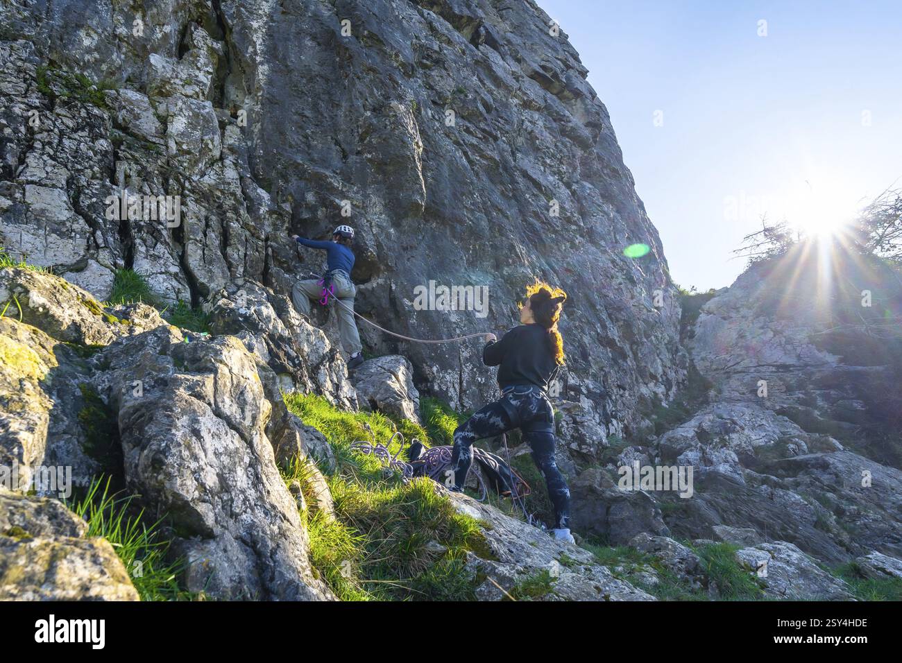 Two female rock climbers scaling a steep cliff face, illuminated by bright sunlight, showcasing ...