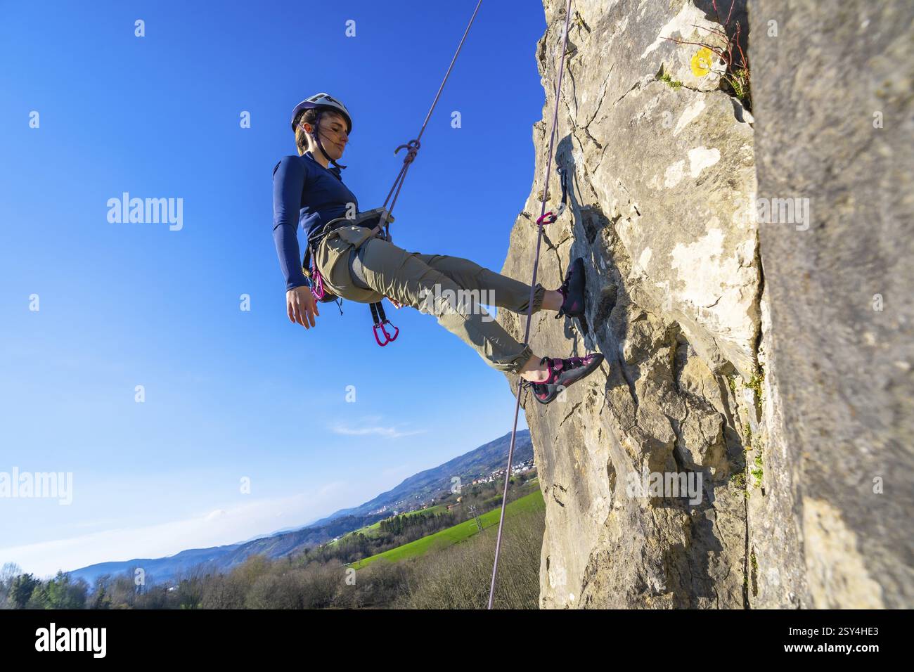 Young woman climber ascending a steep rock face using ropes and climbing gear, enjoying a sunny ...