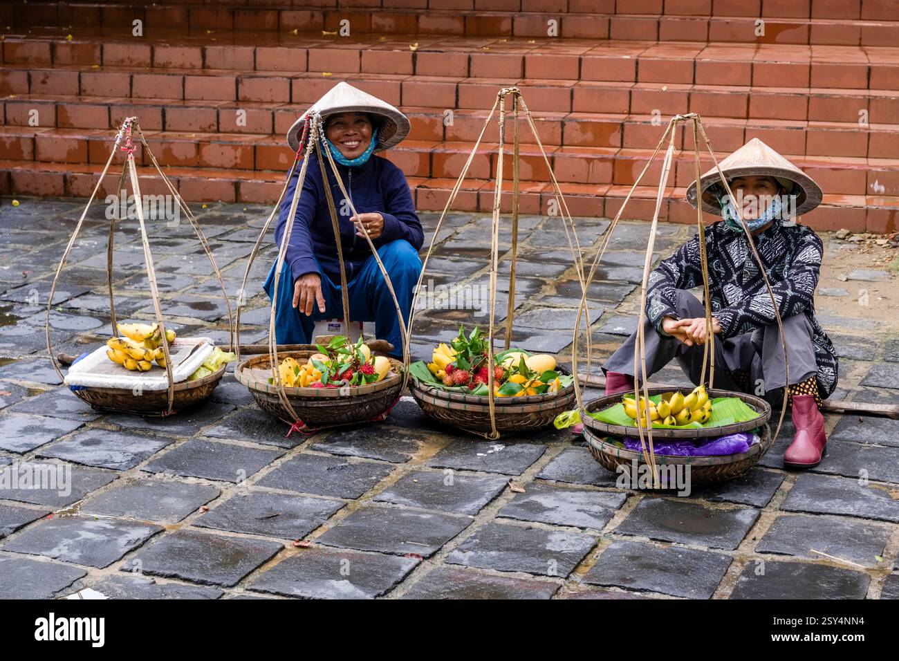 Two women wearing conical hats sell different types of fruit in baskets ...