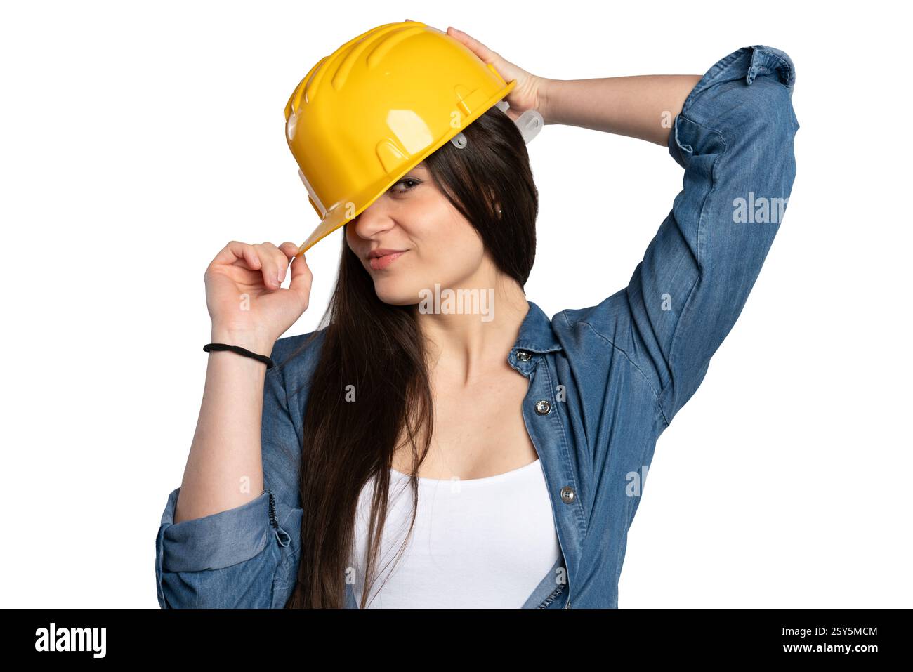 A confident female construction worker playfully adjusting her yellow safety helmet while ...