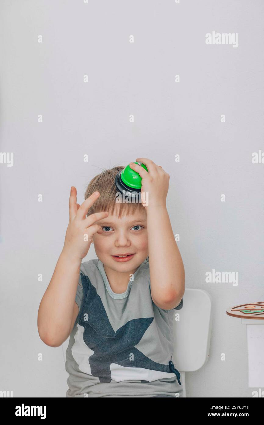 boy in therapy room with green bell balanced on his head. he clasps his ...