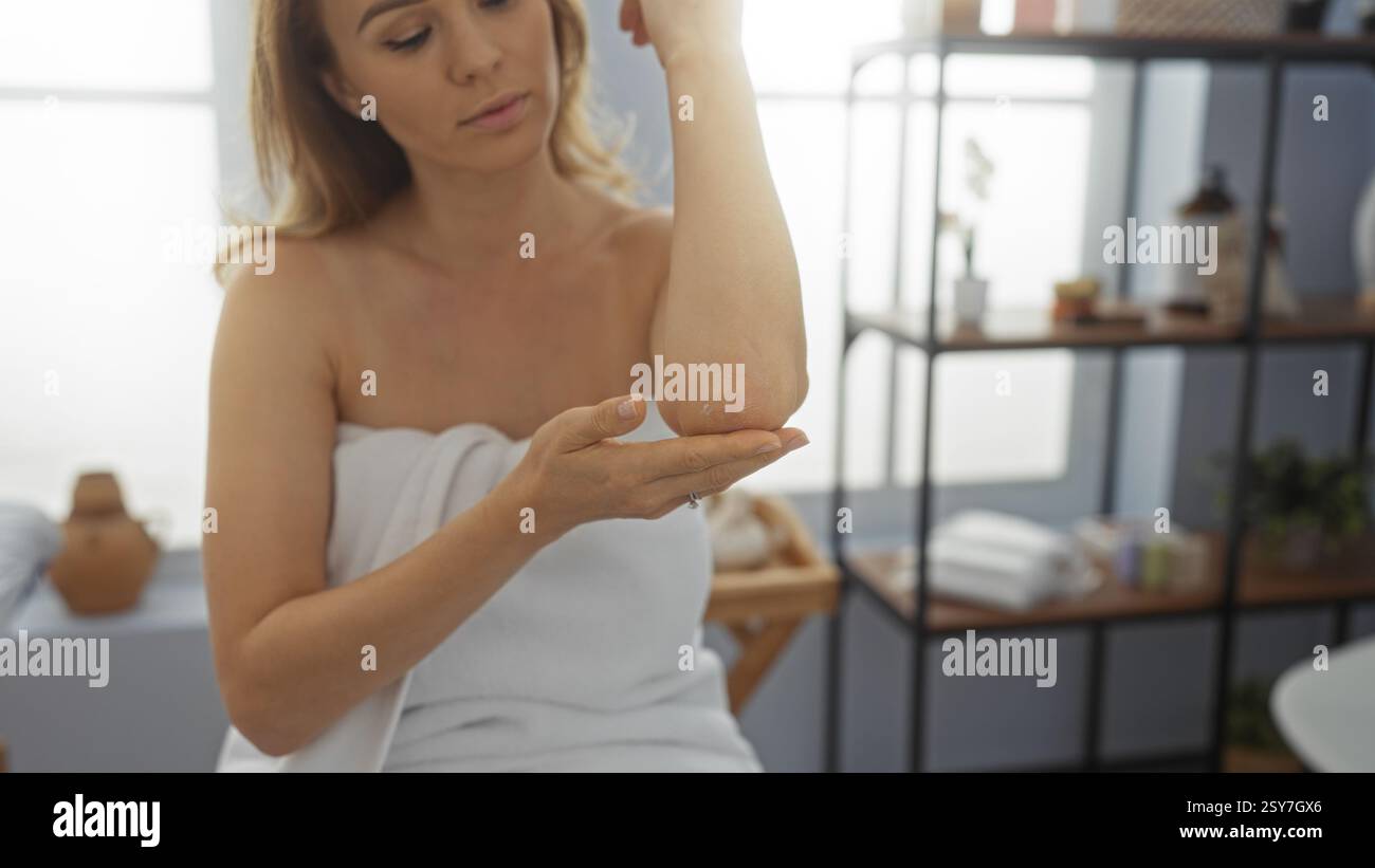 Woman applying skincare on elbow wrapped in towel in tranquil spa room ...